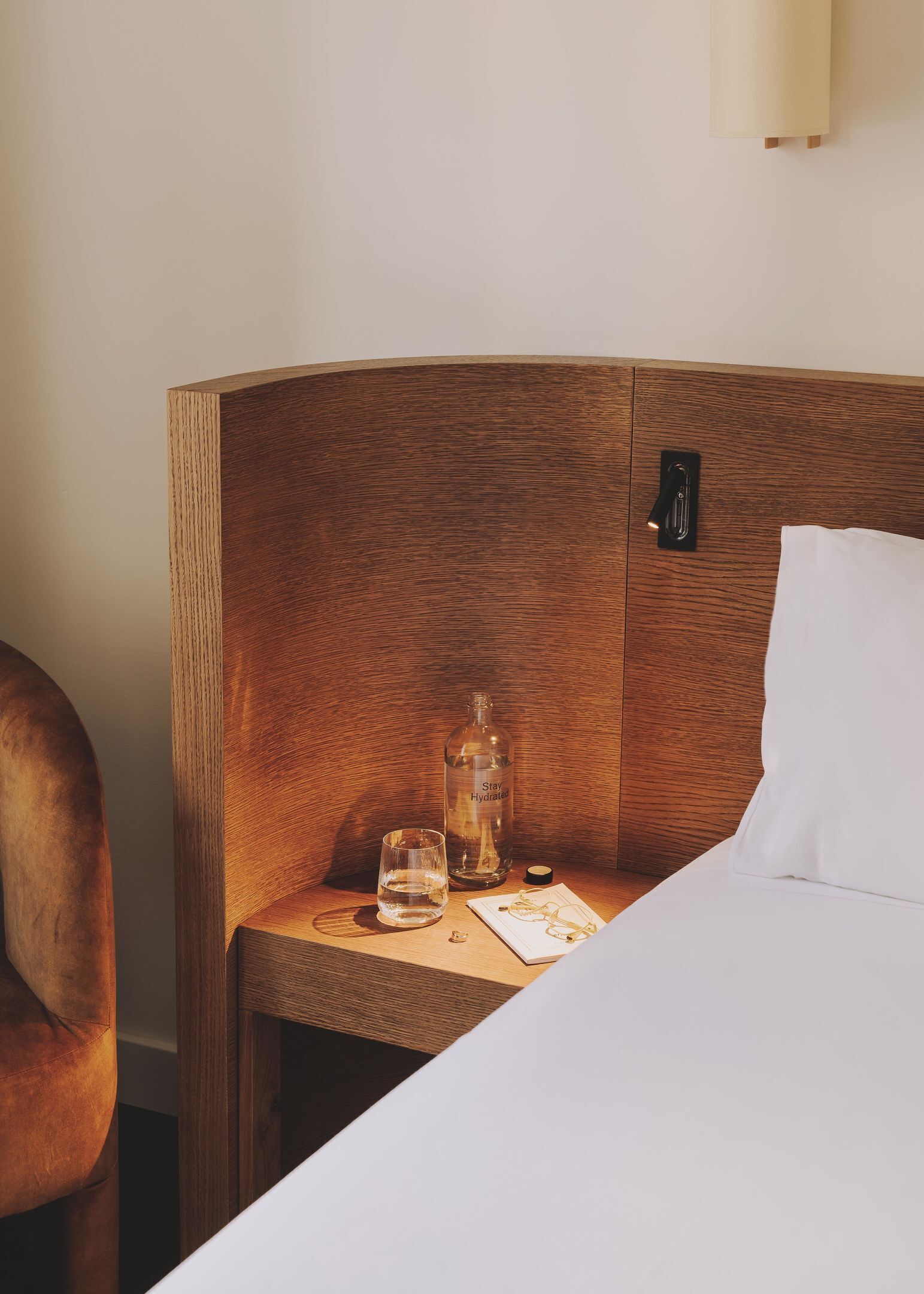 Quarto de hotel com cabeceira de madeira, mesa de cabeceira com uma garrafa de água, um copo e um par de óculos.