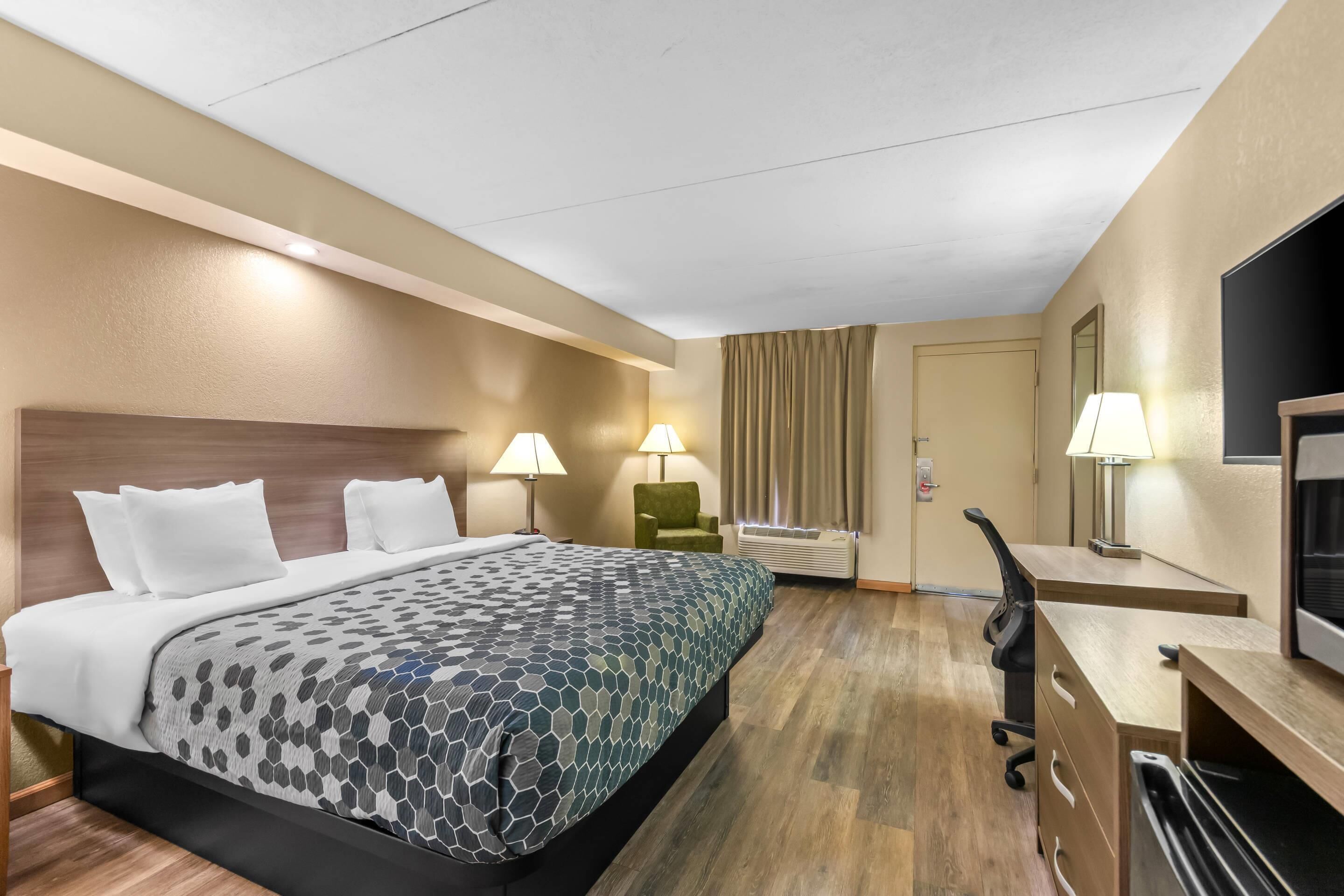 Quarto de hotel com cama, escrivaninha, cadeira, luminárias e televisão.