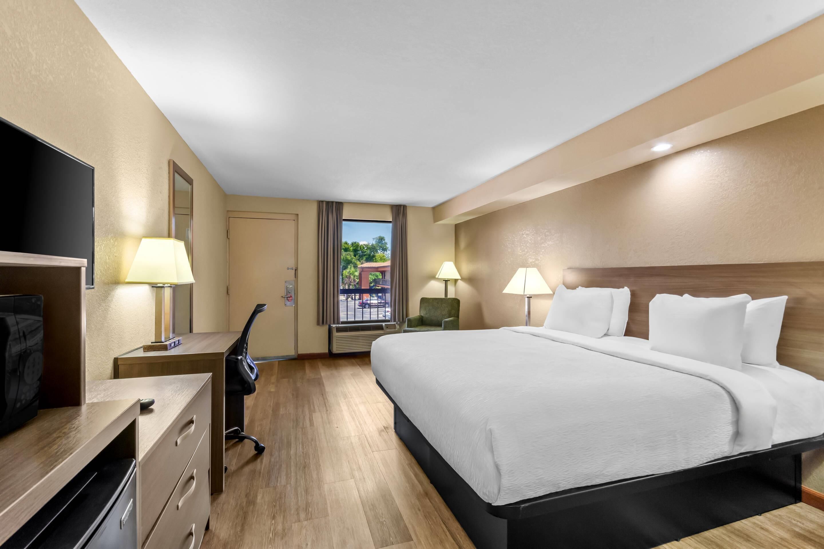 Quarto de hotel com cama, escrivaninha, cadeira, televisão e luminárias.