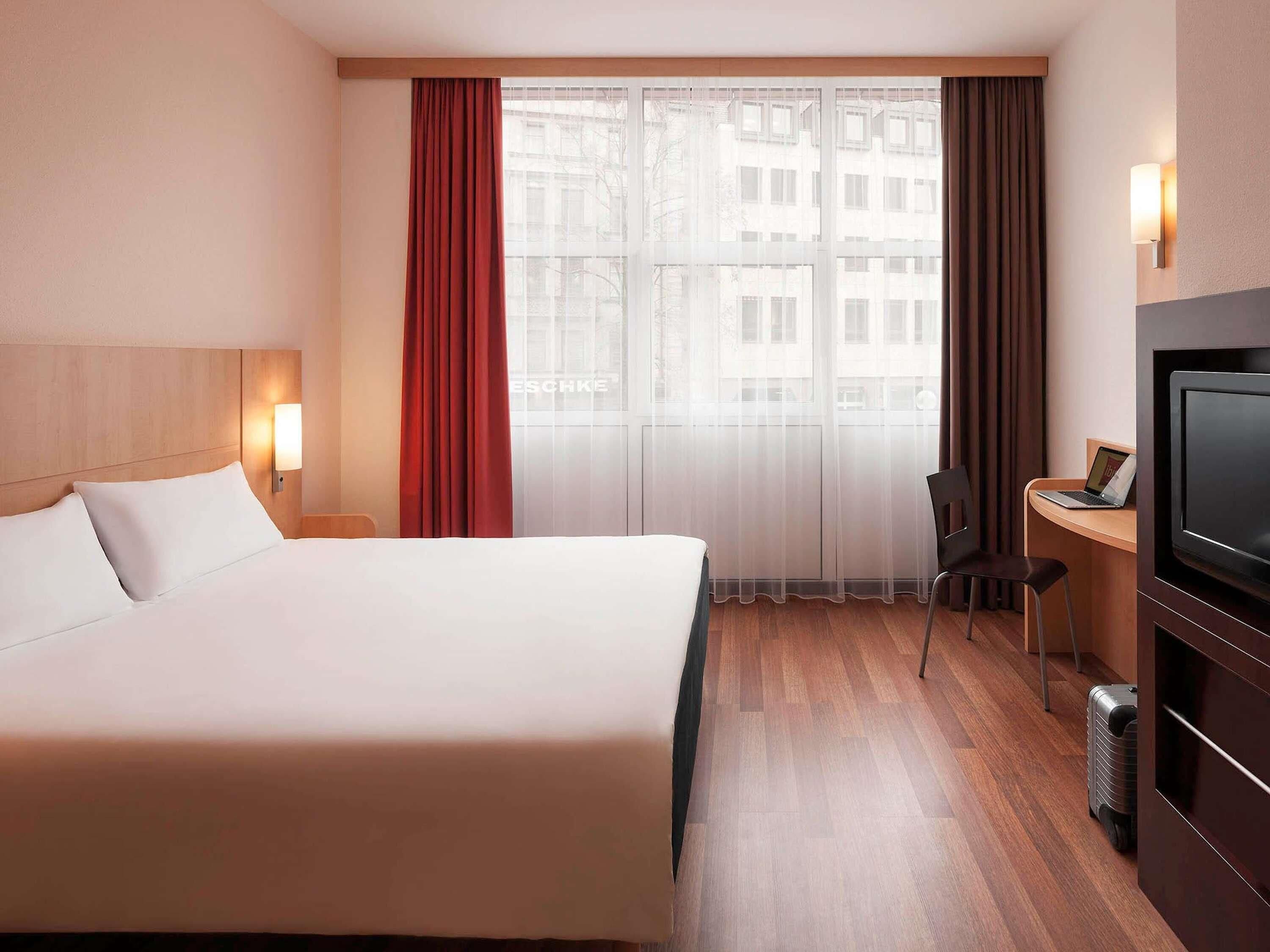 Quarto de hotel com uma cama, uma escrivaninha com um laptop, uma televisão e uma janela com cortinas.