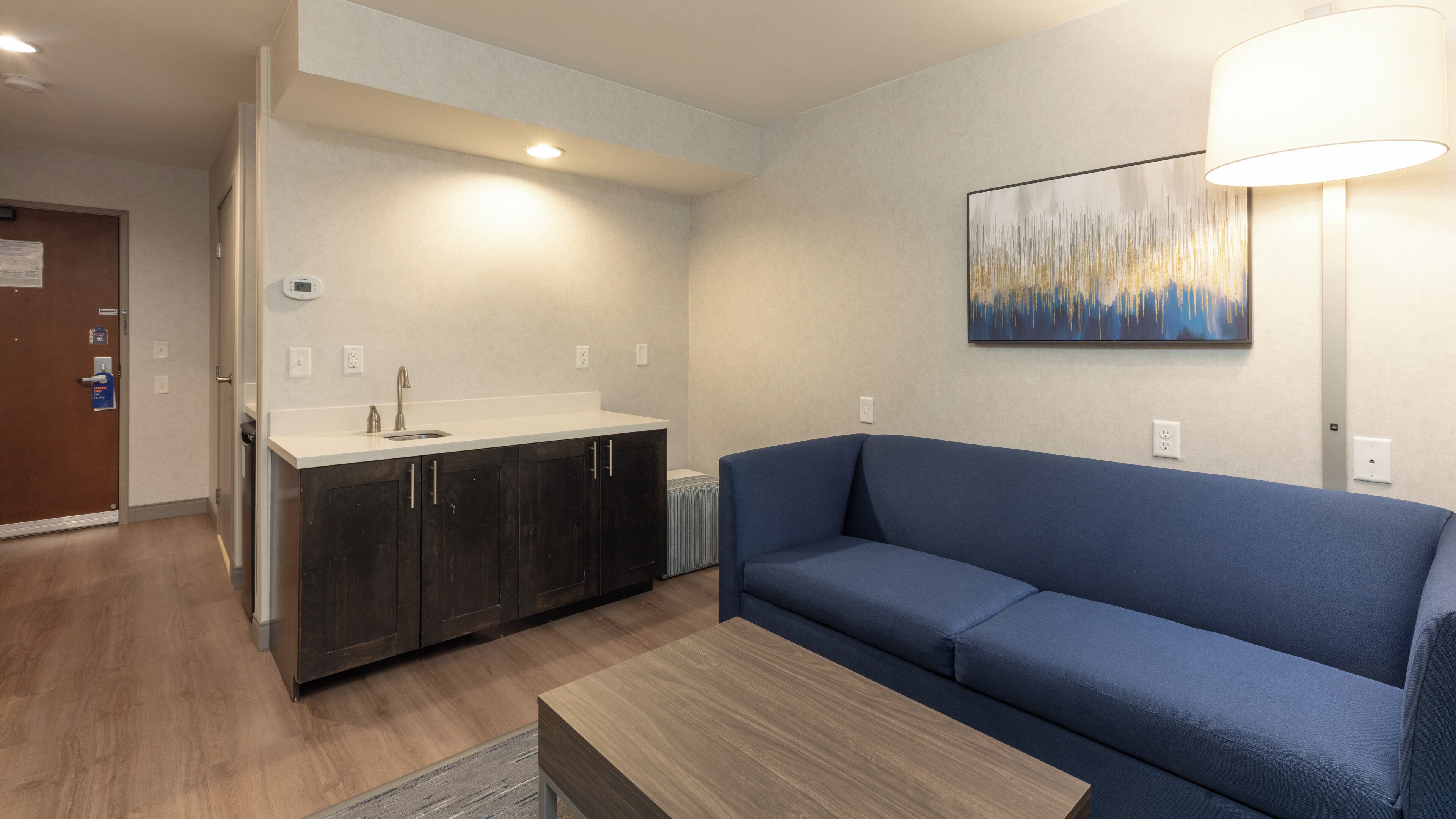 Uma sala de estar moderna com um sofá azul, uma mesa de centro de madeira e uma área de cozinha com armários escuros e bancada branca.