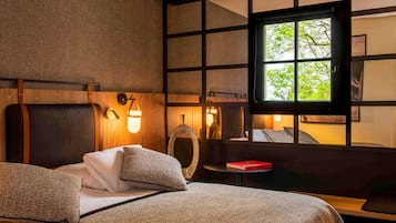 Quarto de hotel com cama, mesa de cabeceira, janela com vista para árvores e luminária fixada na parede.