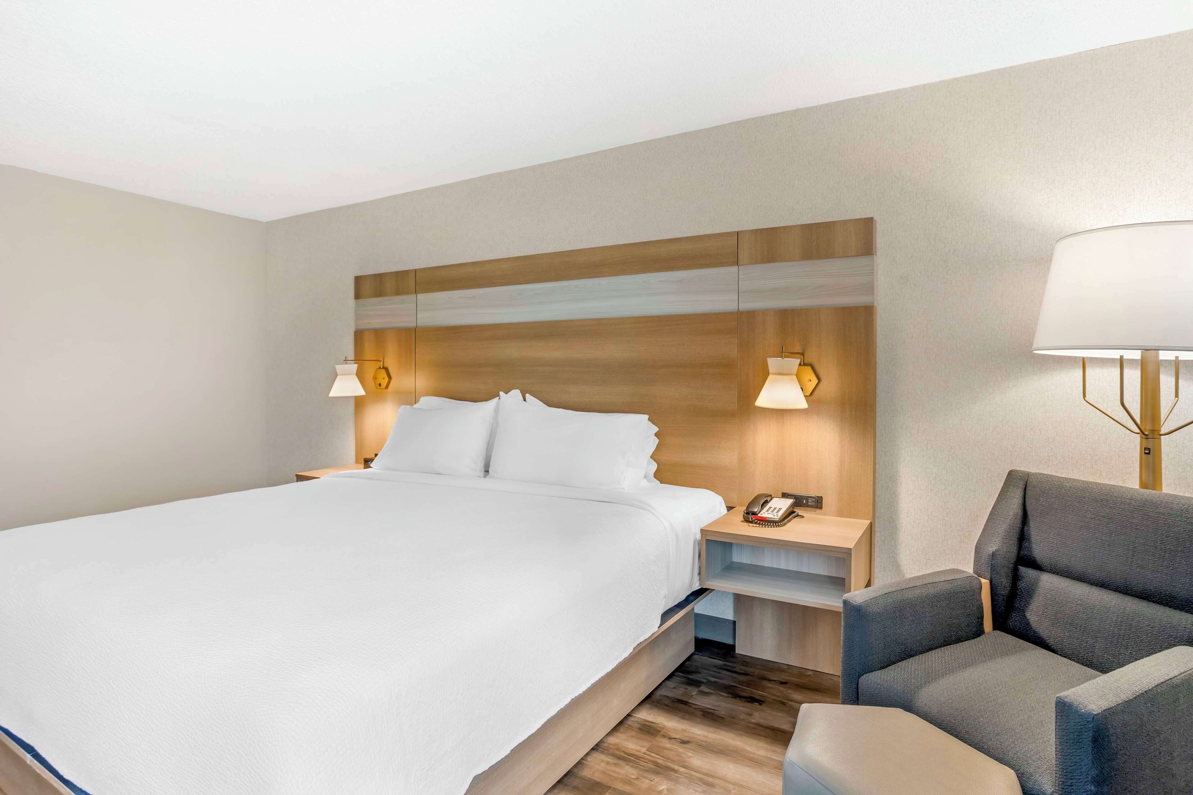 Quarto de hotel com uma cama grande, mesas de cabeceira com luminárias, uma cadeira e uma cabeceira de madeira.