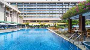 Uma área de piscina de hotel com várias espreguiçadeiras e guarda-sóis, rodeada por um edifício grande com varandas.