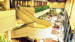 Um átrio de vários níveis com uma escadaria espiral central, plantas internas e uma área de refeições com mesas e cadeiras.