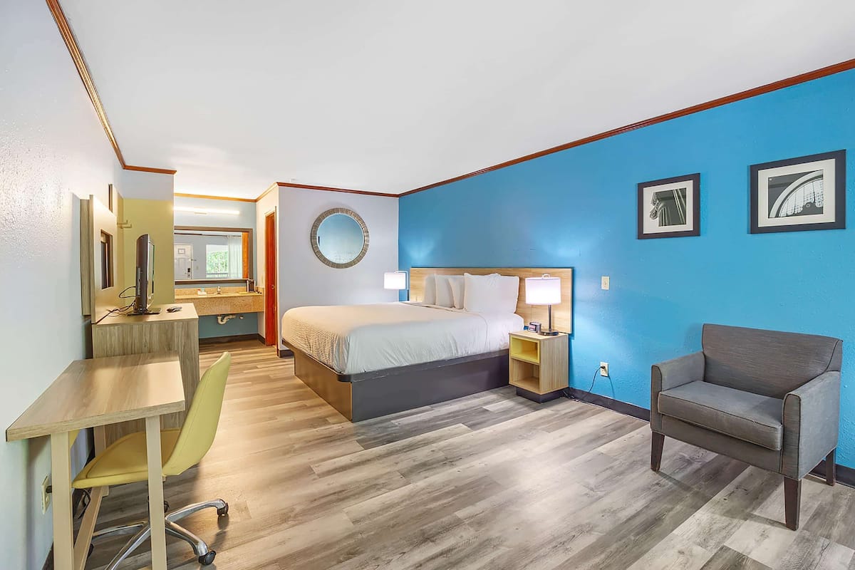 Quarto de hotel com cama, escrivaninha, cadeira, mesa de cabeceira e parede com quadros.