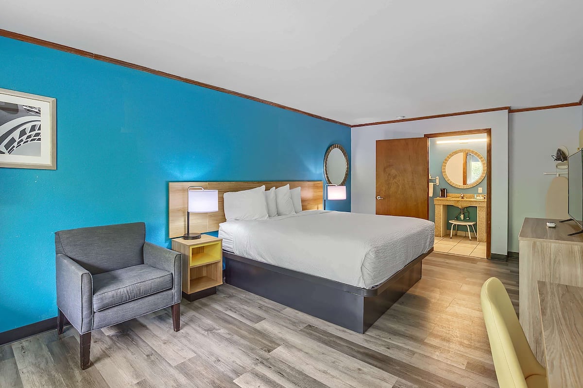Quarto de hotel com uma cama, uma cadeira, uma escrivaninha e um banheiro visível através de uma porta aberta.