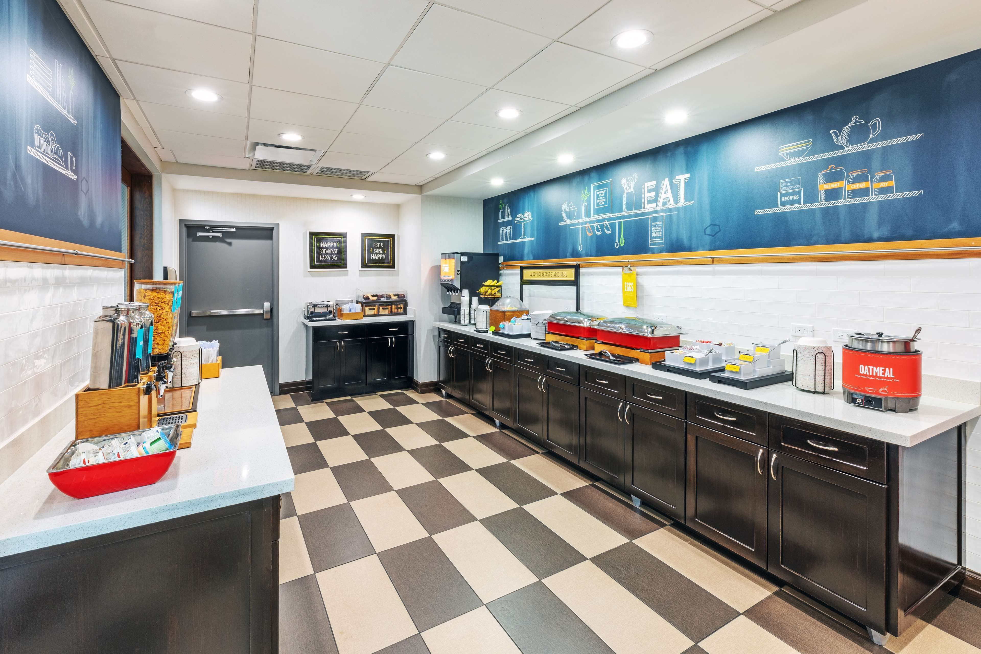 Uma cozinha moderna com piso xadrez, balcão com diversos alimentos e parede de lousa com desenhos relacionados à culinária.