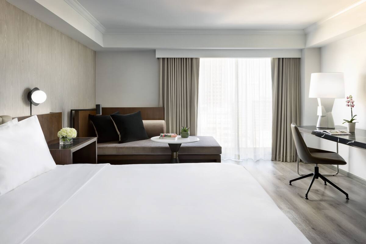 Quarto de hotel com cama, escrivaninha, cadeira, mesa redonda, abajur e janela com cortinas.
