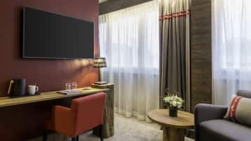 Quarto de hotel com televisão de tela plana na parede, uma secretária de madeira com cadeira, uma mesa redonda pequena com um vaso de flores e um sofá com travesseiro decorativo.