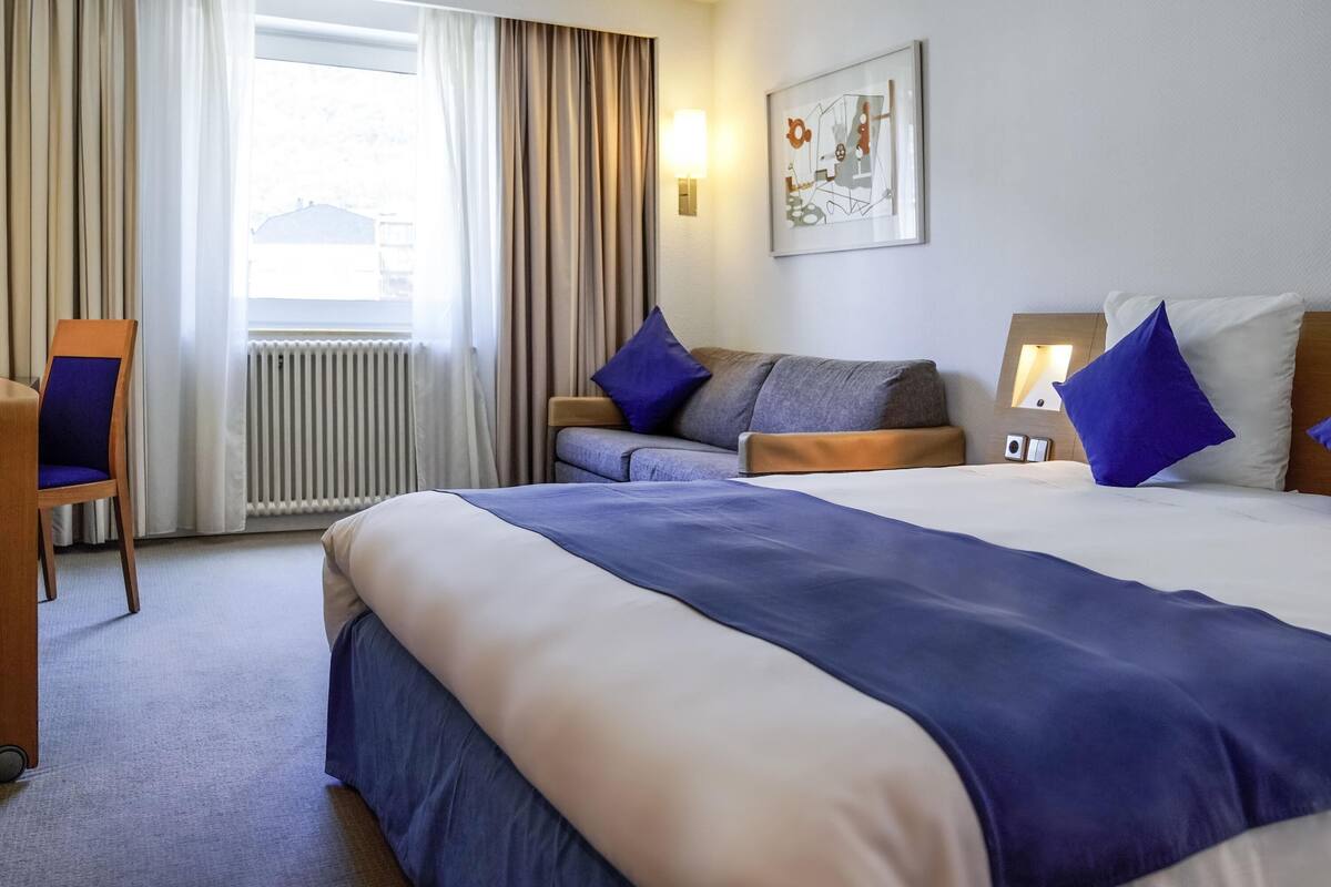 Quarto de hotel com uma cama grande, um sofá, uma cadeira e uma janela com cortinas.