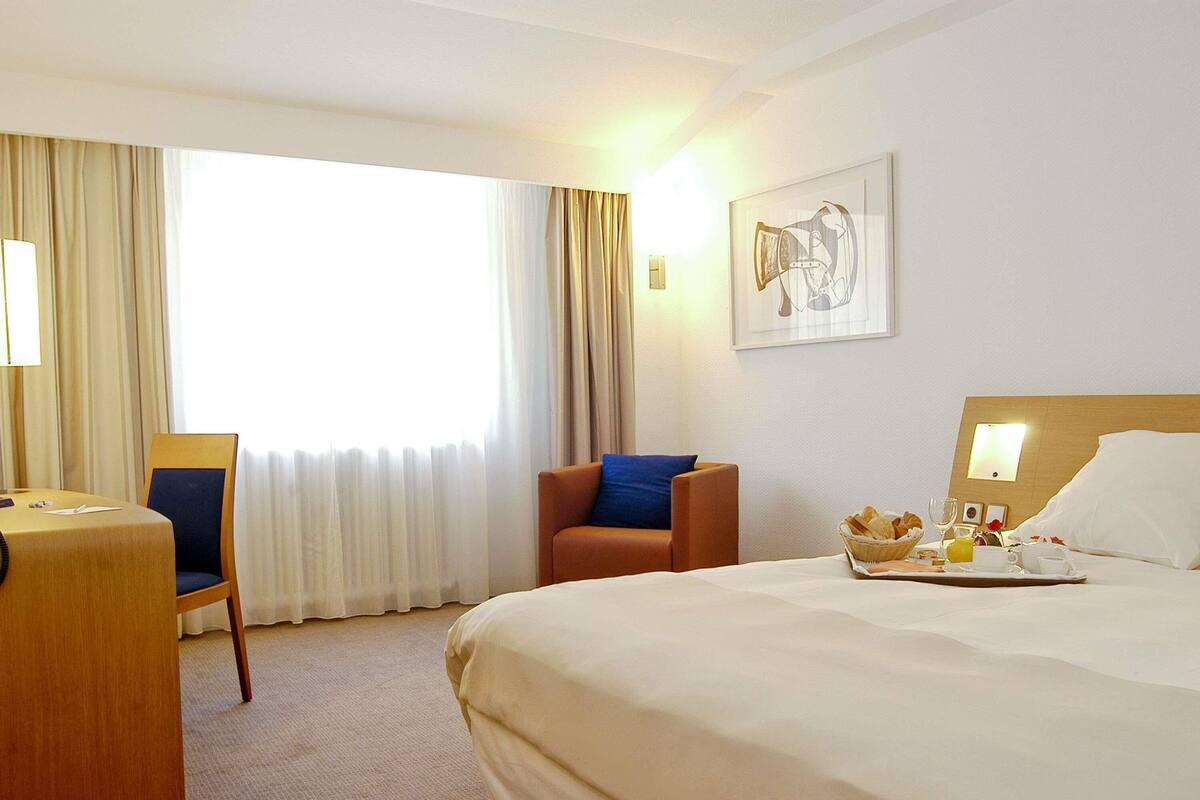 Quarto de hotel com uma cama, uma mesa com cadeira, uma janela com cortinas e uma bandeja de café da manhã sobre a cama.