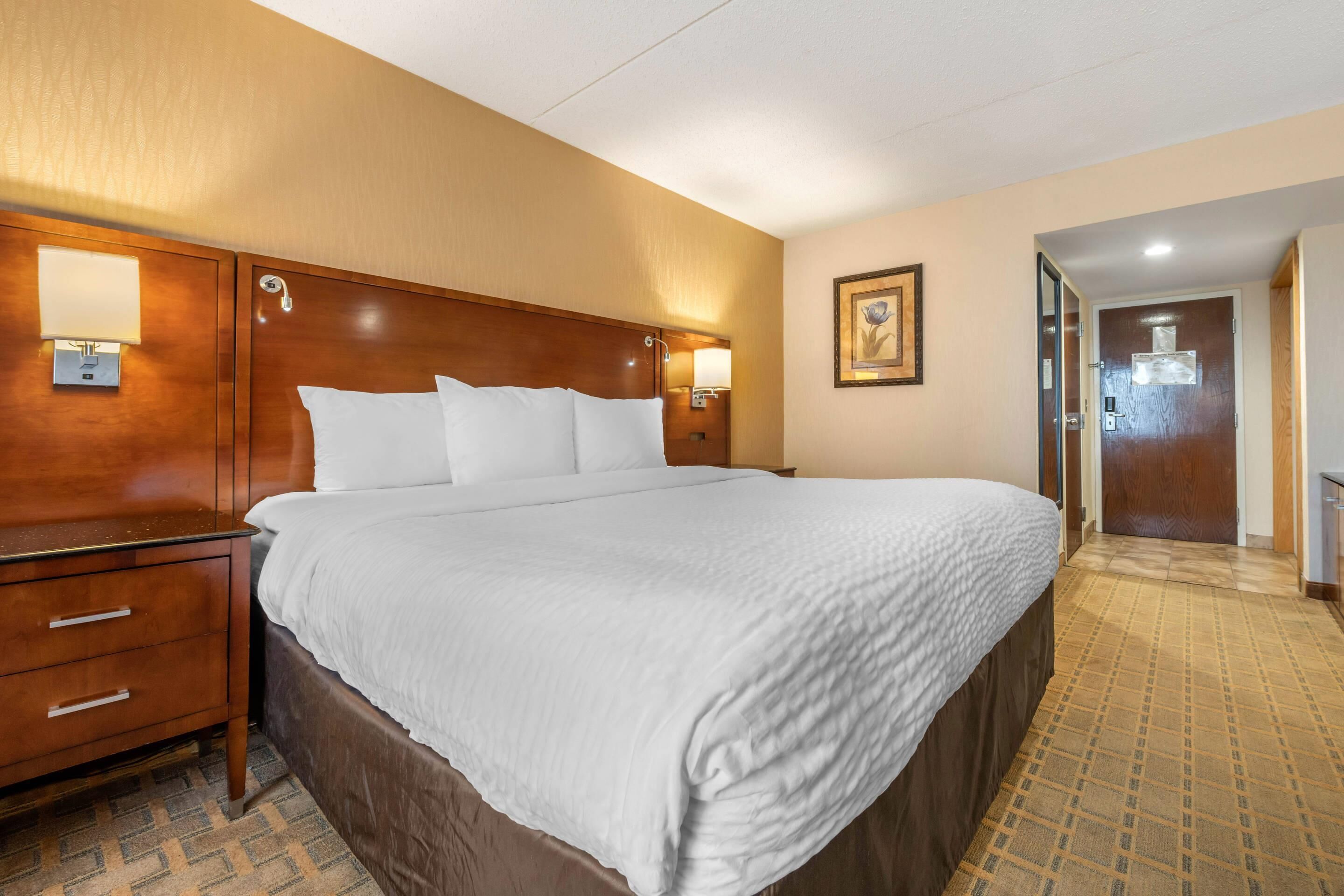 Quarto de hotel com uma cama grande, cabeceira de madeira, luminárias laterais, mesa de cabeceira e um tapete estampado.