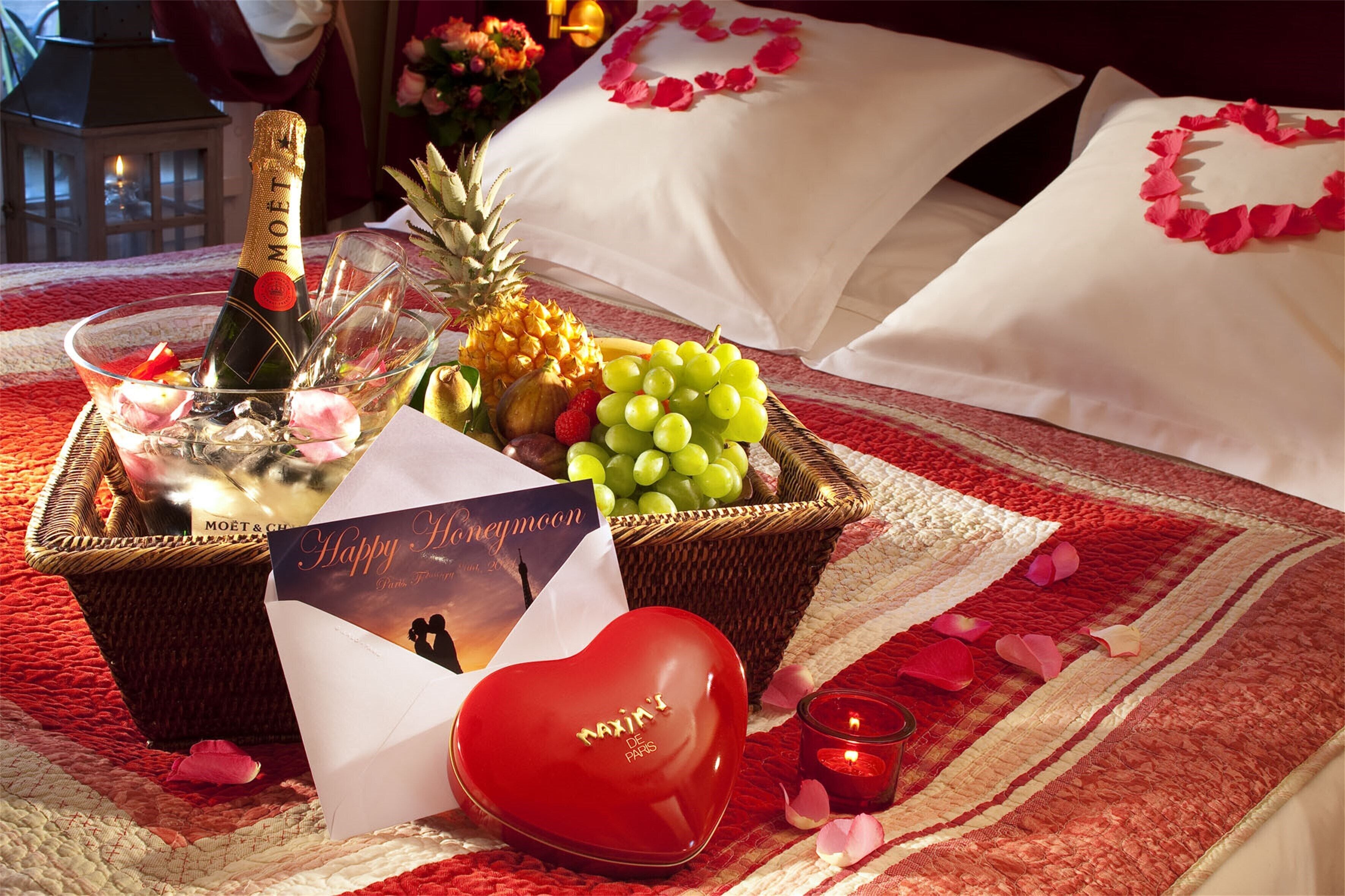 Ambiente romântico com uma garrafa de Moët champagne, uma cesta de frutas, uma caixa em formato de coração vermelha e uma vela sobre uma cama de pétalas de rosa.