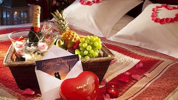 Ambiente romântico com uma garrafa de Moët champagne, uma cesta de frutas, uma caixa em formato de coração vermelha e uma vela sobre uma cama de pétalas de rosa.