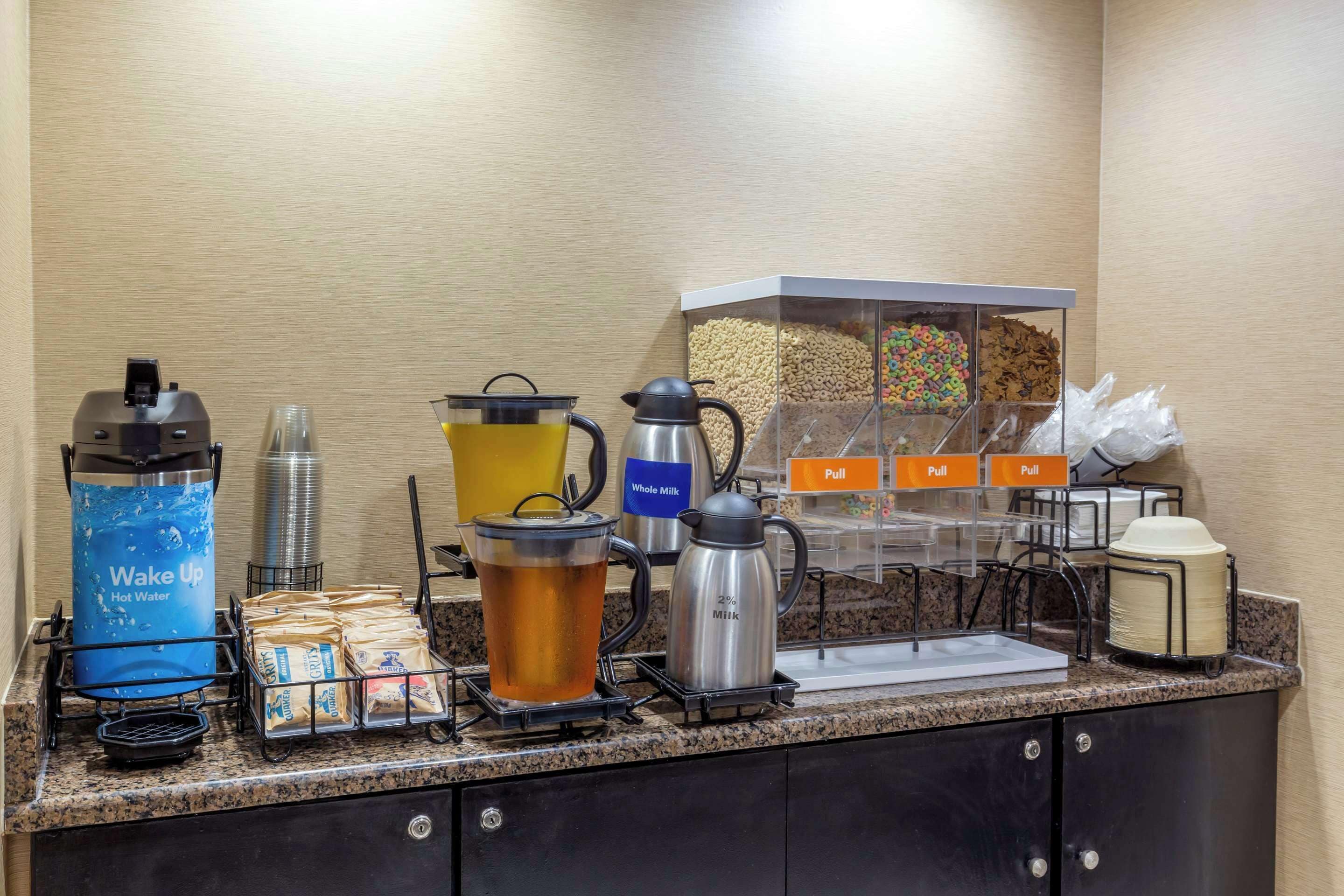 Um buffet de café da manhã de hotel com máquina de café, chaleira, jarra de café, dispensador de água e um recipiente com cereal.