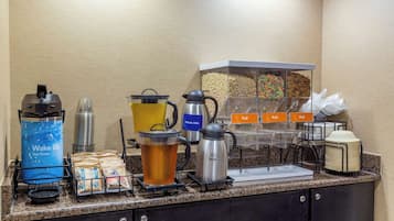 Um buffet de café da manhã de hotel com máquina de café, chaleira, jarra de café, dispensador de água e um recipiente com cereal.