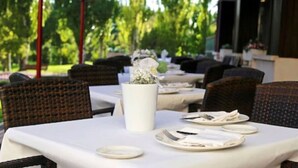 Uma mesa posta para refeições ao ar livre, com cadeiras de vime, toalha de mesa branca e um vaso com flores.