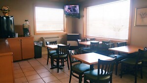 Restaurante com várias mesas e cadeiras, uma máquina de café, uma televisão na parede e uma janela com persianas.