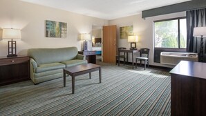 Quarto de hotel com sofá, mesa pequena, escrivaninha, cadeira, luminária e janela com cortinas.