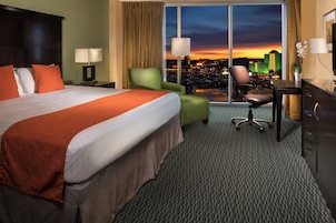 Quarto de hotel com uma cama grande, uma escrivaninha, uma poltrona verde e vista para a paisagem urbana ao pôr do sol.