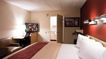 Quarto de hotel com cama, escrivaninha com televisão, cadeira, porta e luminária fixada na parede.