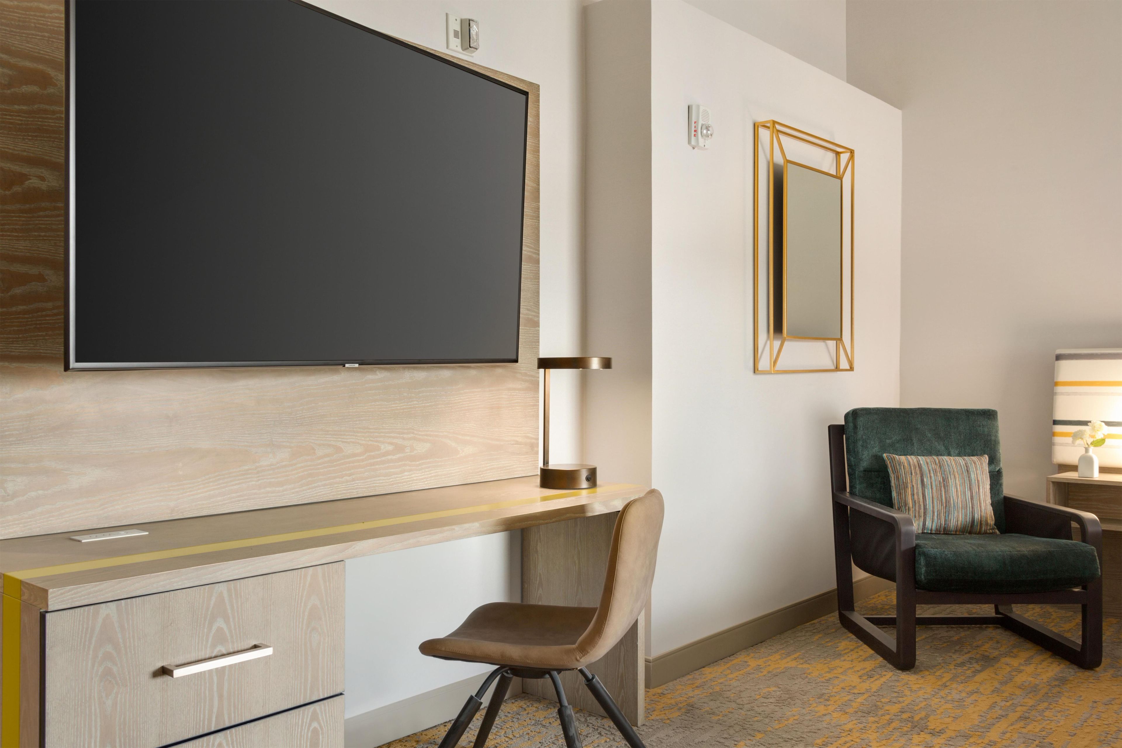 Quarto de hotel com uma televisão de tela plana grande fixada em uma parede de madeira, uma escrivaninha com gavetas, uma poltrona estofada, uma poltrona verde e uma mesinha de cabeceira com abajur.