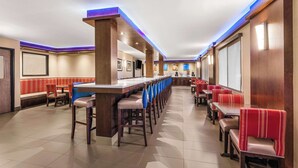 Uma extensa área de bar com banquetas e mesas altas, um balcão e um espaço de estar com cadeiras estofadas vermelhas.