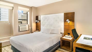 Quarto de hotel com uma cama grande, uma escrivaninha com cadeira, uma luminária na parede e uma janela com persianas.