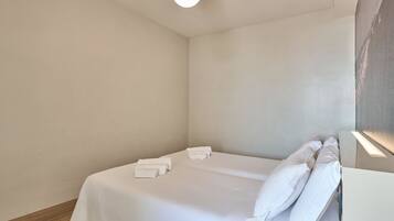 Quarto de hotel minimalista com uma cama simples, cuidadosamente arrumada com roupa de cama e almofadas brancas, piso de madeira e um design limpo e descomplicado.