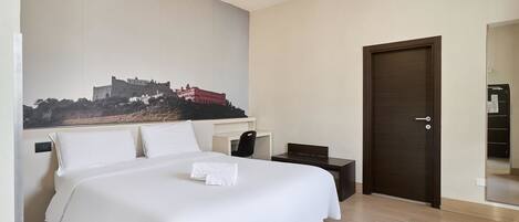 Quarto de hotel com cama, escrivaninha, cadeira, porta e luminária na parede.