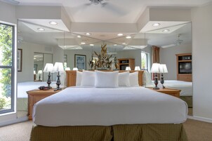 Uma cama grande com lençóis brancos, uma mesa de cabeceira com abajur, um espelho sobre a cama e um ventilador de teto.