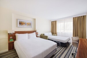 Quarto de hotel com duas camas, uma escrivaninha, uma cadeira, um quadro na parede e uma janela com cortinas.
