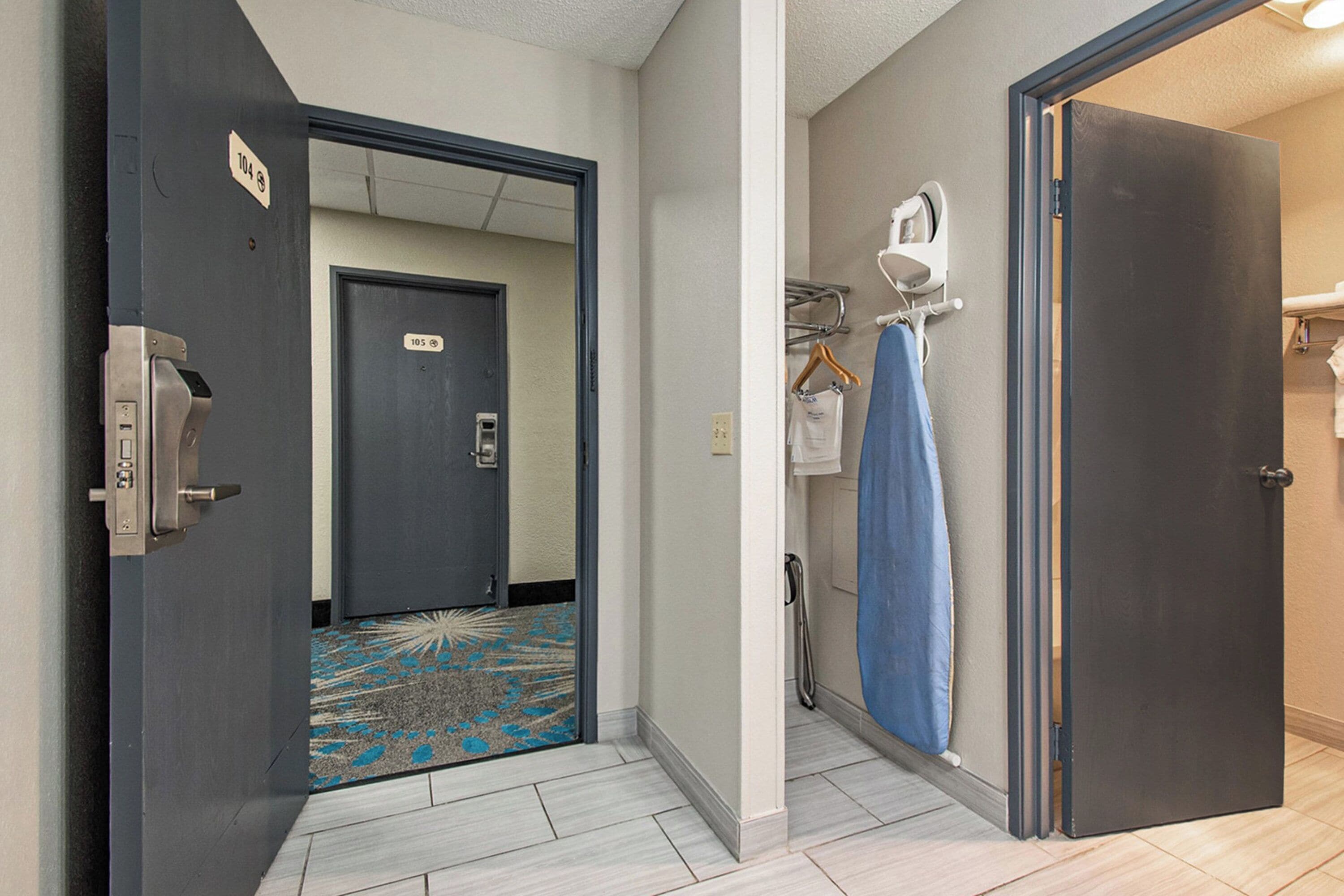 Corredor de hotel com uma porta identificada com o número "104" dando acesso a um quarto, uma toalha azul pendurada em um suporte e uma área de chuveiro com um porta-toalhas.