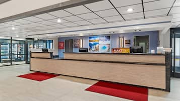 Uma área de recepção de hotel com design moderno, incluindo um balcão de atendimento, uma tela digital e um grande letreiro de boas-vindas.