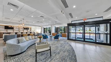 Um saguão moderno de hotel com um sofá curvo, uma mesa de centro e cadeiras azuis. Há uma área de cozinha com uma TV e uma mesa de jantar. O saguão possui uma grande porta de vidro que dá para o exterior.