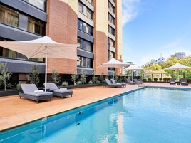 Uma área à beira da piscina com espreguiçadeiras e guarda-sóis, piscina azul límpida e um edifício com fachada de tijolos.