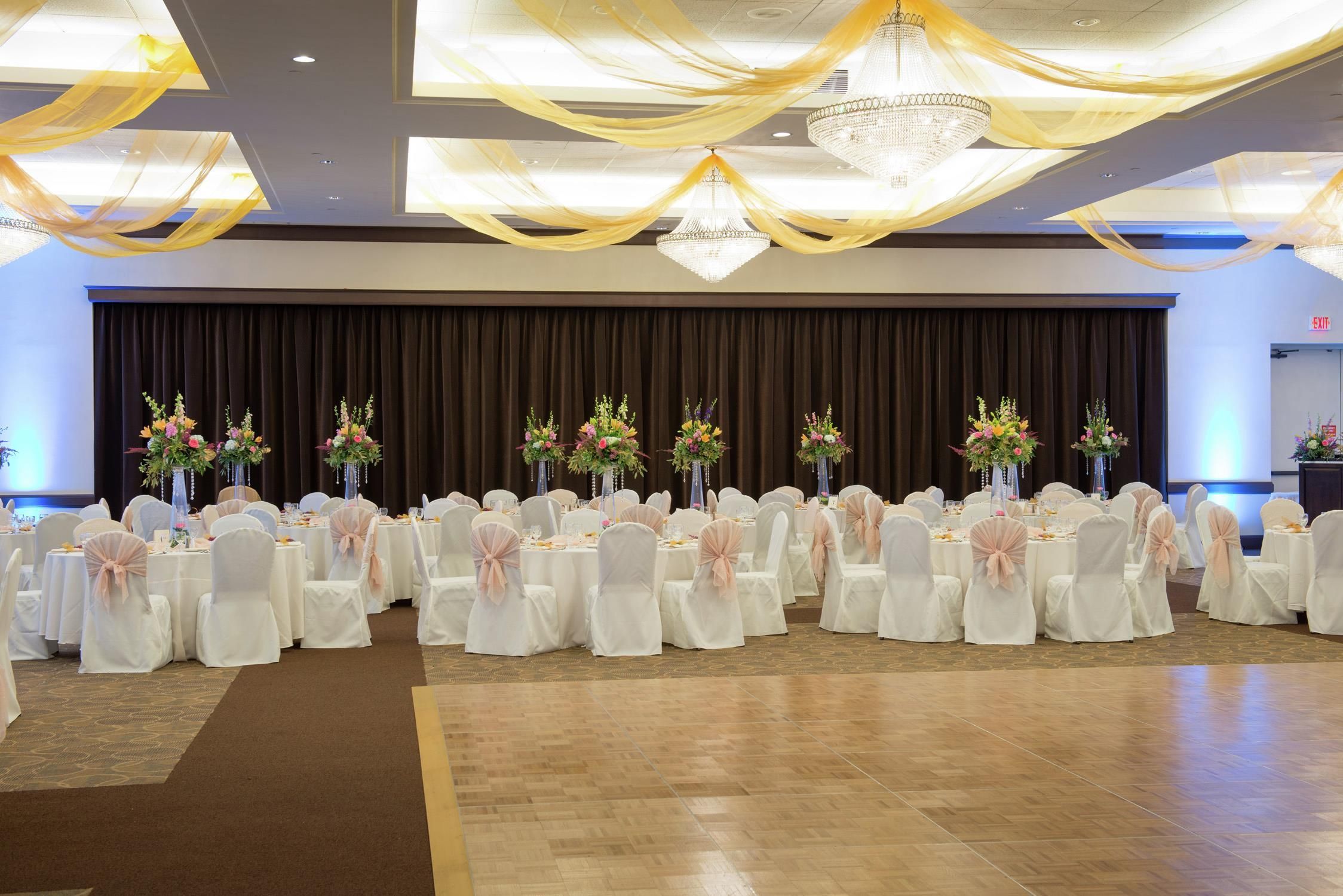 Uma sala de banquetes com mesas redondas, cadeiras brancas e arranjos florais.