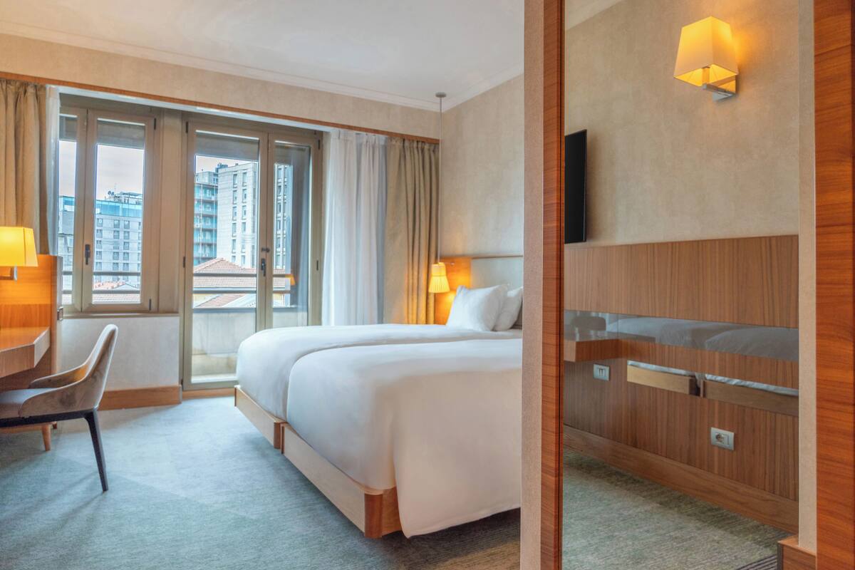 Quarto de hotel com uma cama grande, uma mesa, uma cadeira, uma TV e uma janela com cortinas.