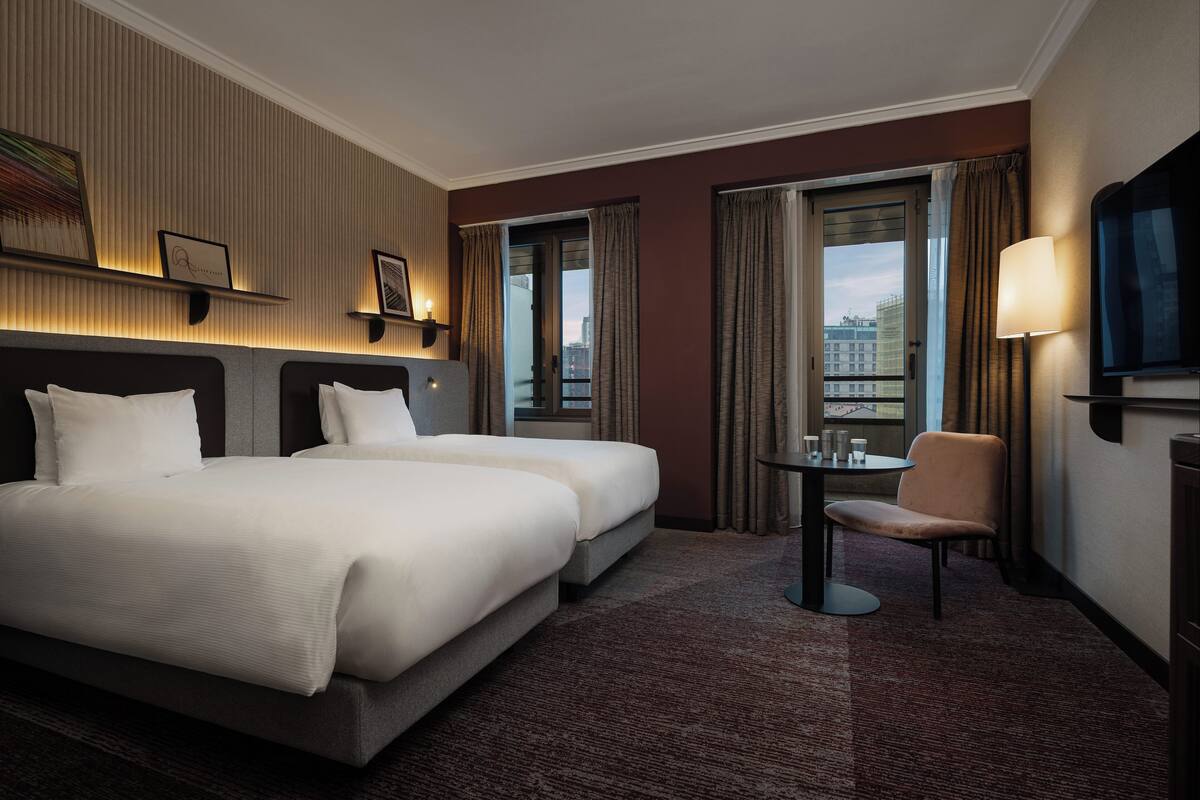 Quarto de hotel com duas camas, uma escrivaninha, uma cadeira, uma televisão e uma janela com cortinas.