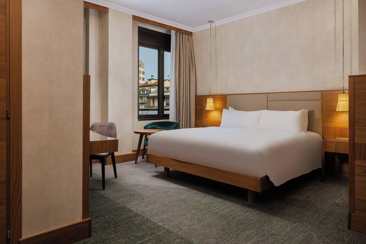 Quarto de hotel com uma cama grande, uma escrivaninha com cadeira, uma janela com vista para edifícios e um abajur na mesa de cabeceira.