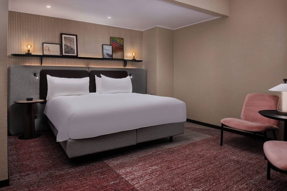 Quarto de hotel com uma cama grande, cabeceira com iluminação embutida, mesa pequena com abajur, uma cadeira e tapete vermelho.