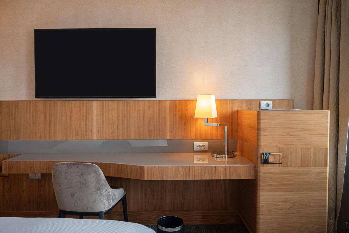 Quarto de hotel com escrivaninha de madeira, cadeira, televisão de tela plana na parede e mesa de cabeceira com abajur.