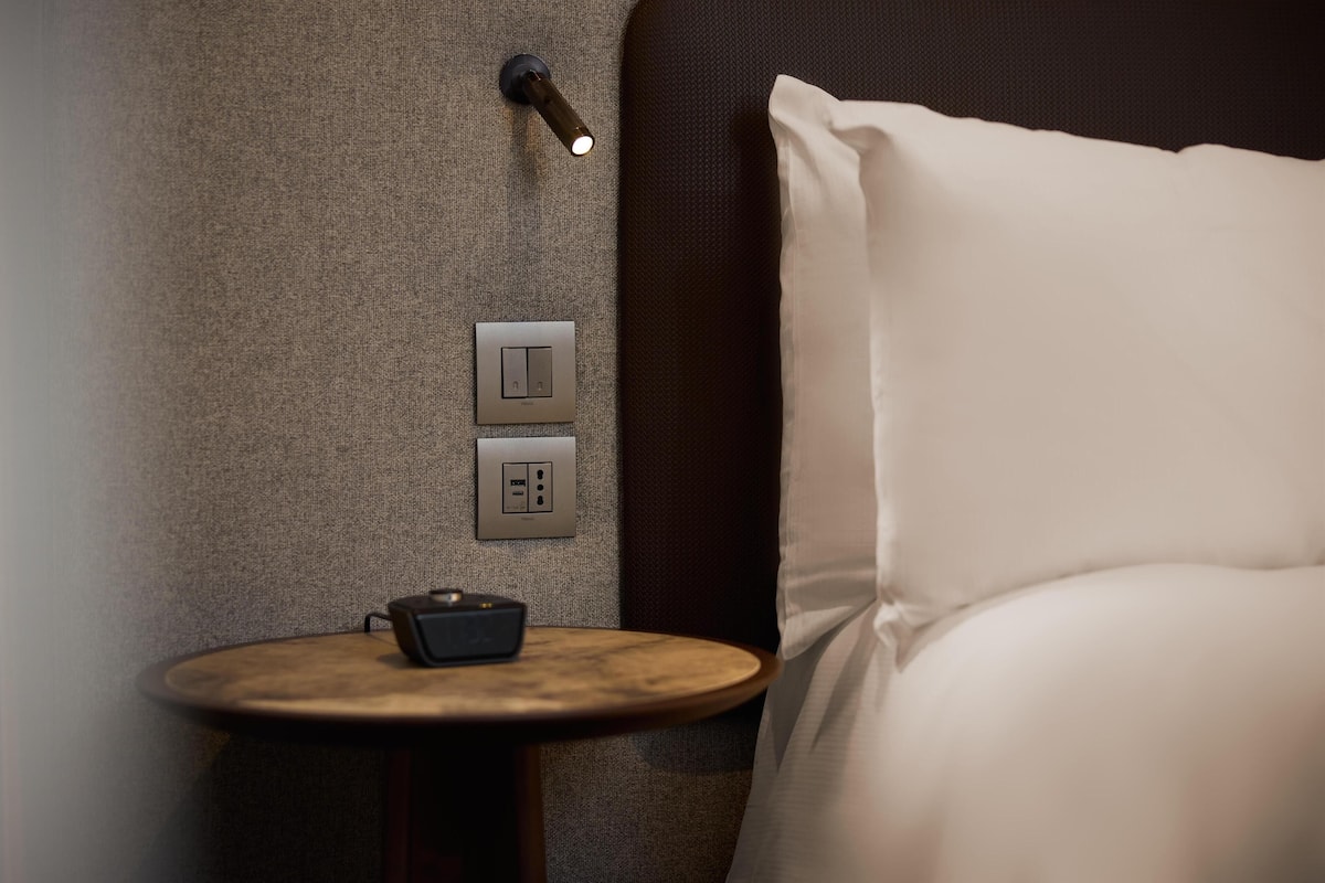 Uma cama bem arrumada com roupa de cama branca, uma pequena mesa redonda com um controle remoto e duas interruptores fixados na parede.
