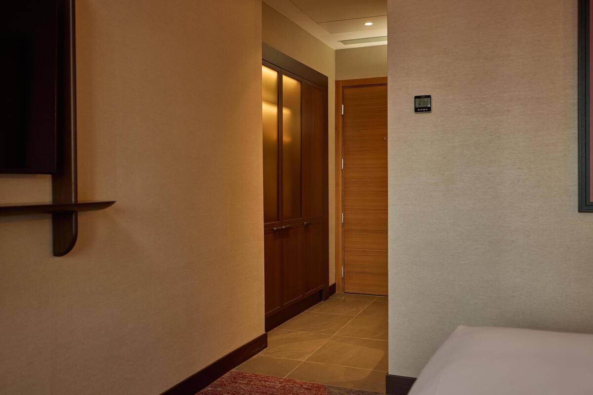 Corredor de hotel com porta de madeira, relógio digital e cama com colcha branca.