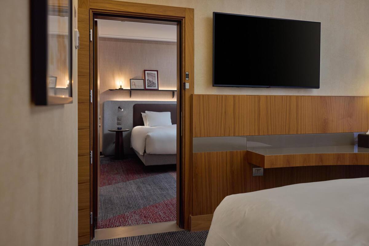 Quarto de hotel com cama, televisão presa à parede, mesa de cabeceira e porta que dá para um banheiro.