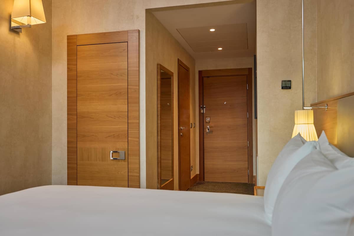 Quarto de hotel com cama, guarda-roupa de madeira e porta que dá para outro cômodo.