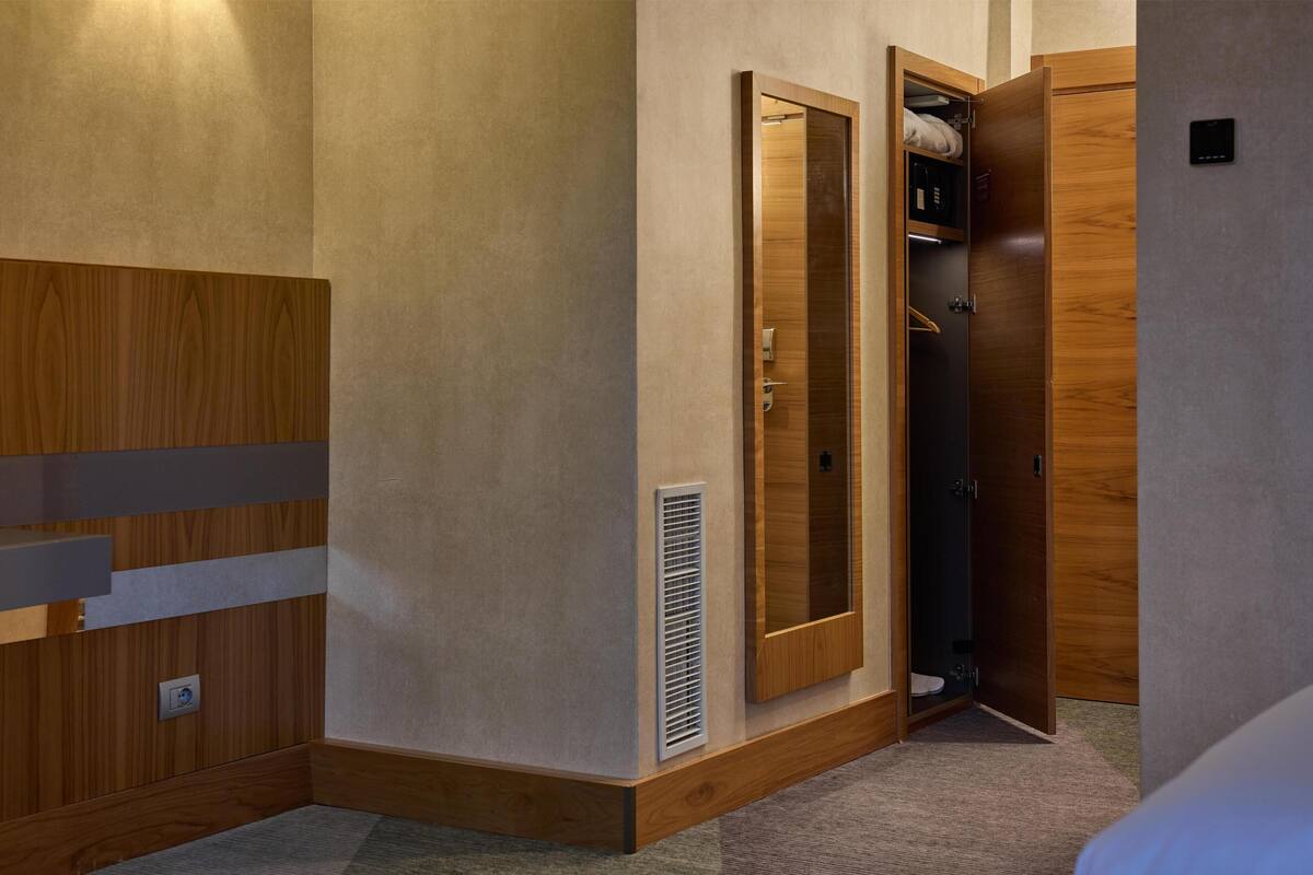 Quarto de hotel com um guarda-roupa de madeira, um espelho, um respiradouro e chão coberto de carpete.