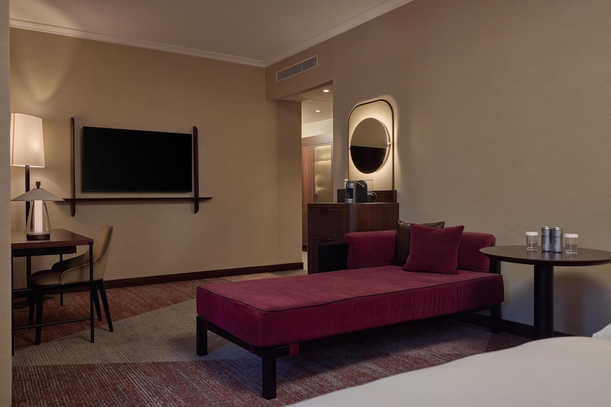 Quarto de hotel com uma cama, uma escrivaninha com abajur, uma televisão, um espelho e uma mesinha com latas.