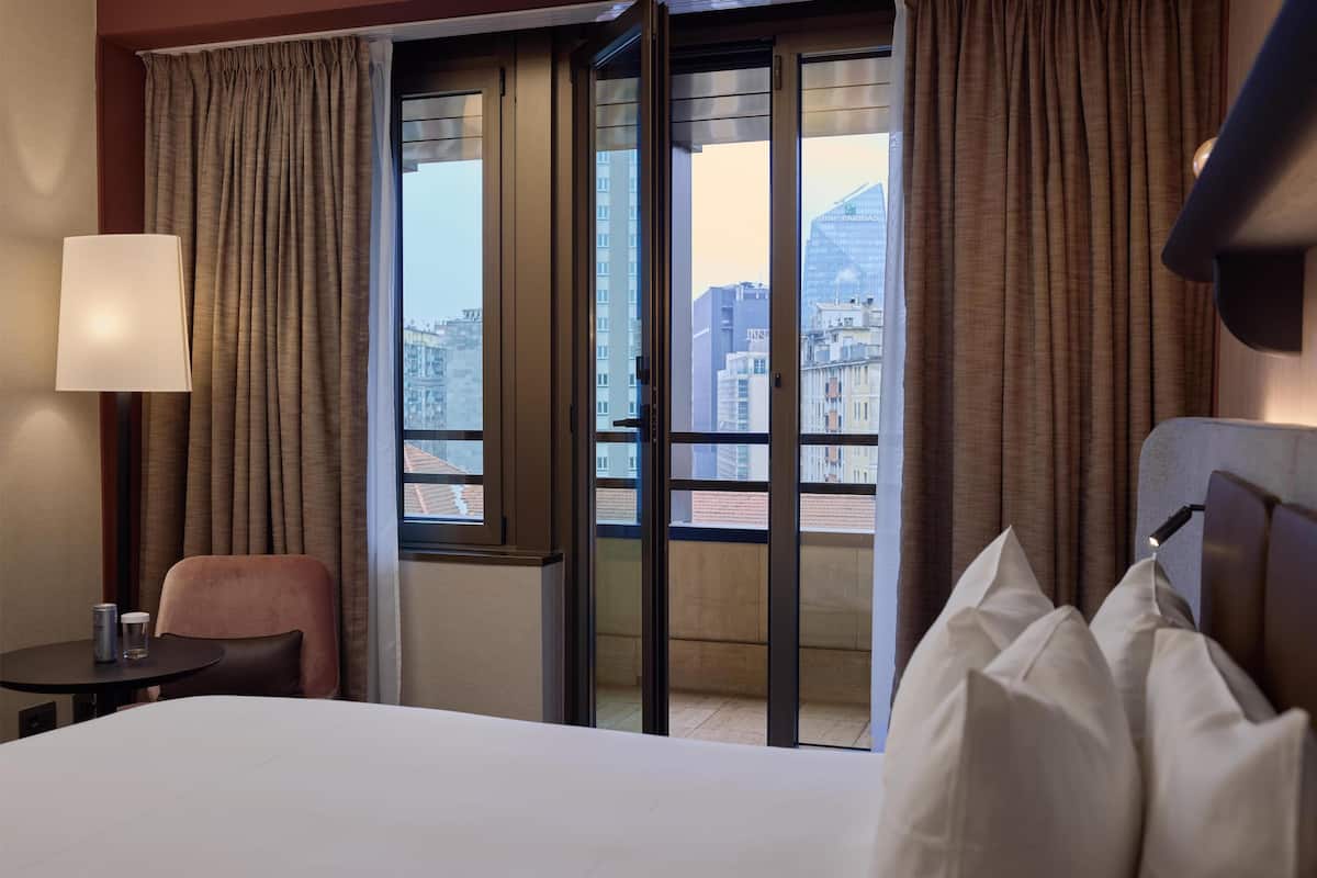 Quarto de hotel com cama, uma cadeira, mesa com bebida e vista para edifícios pela janela.